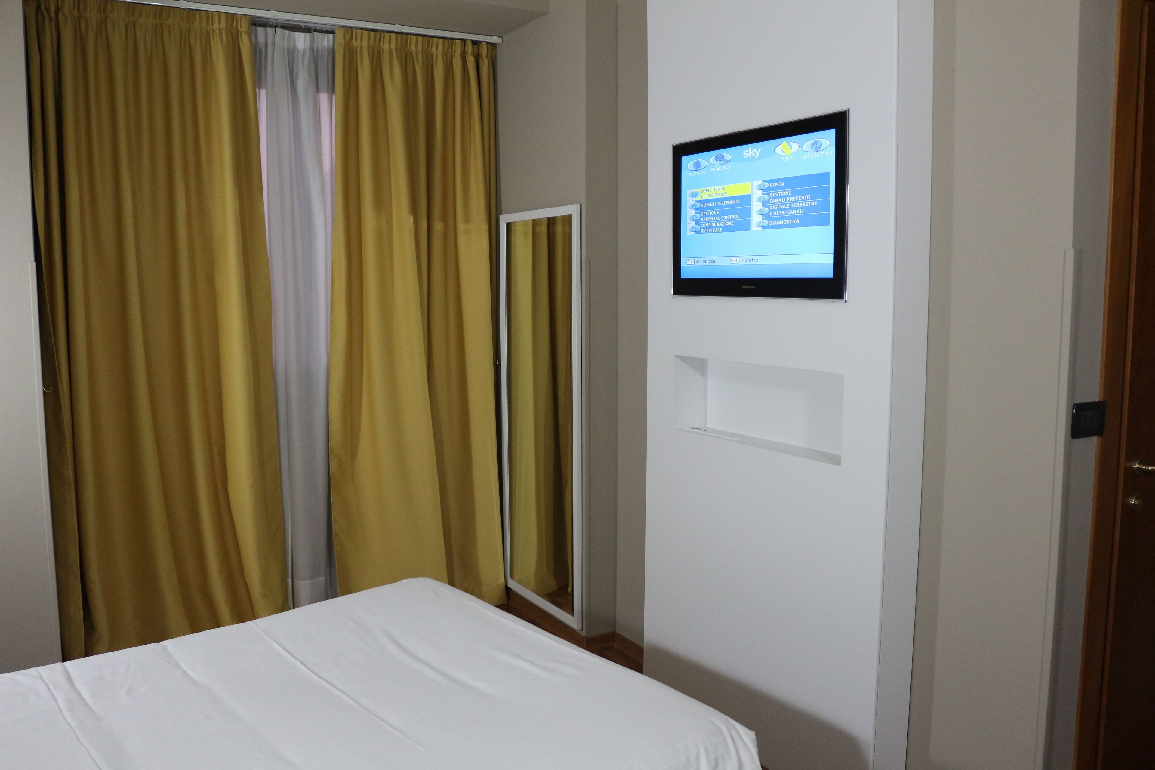 Quarto de hotel com uma cama, televisão fixada na parede e janela com cortinas amarelas.