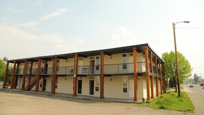 Um motel de dois andares com uma varanda no segundo andar, uma escada que leva à entrada e um estacionamento em frente.