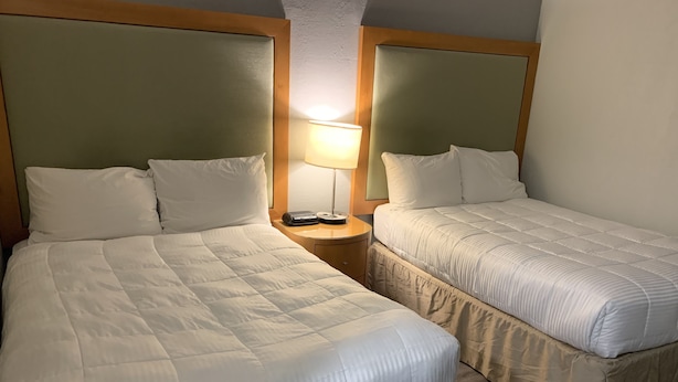 Quarto de hotel com duas camas, cada uma com roupa de cama branca e cabeceiras. Há uma mesa de cabeceira com abajur entre as camas.