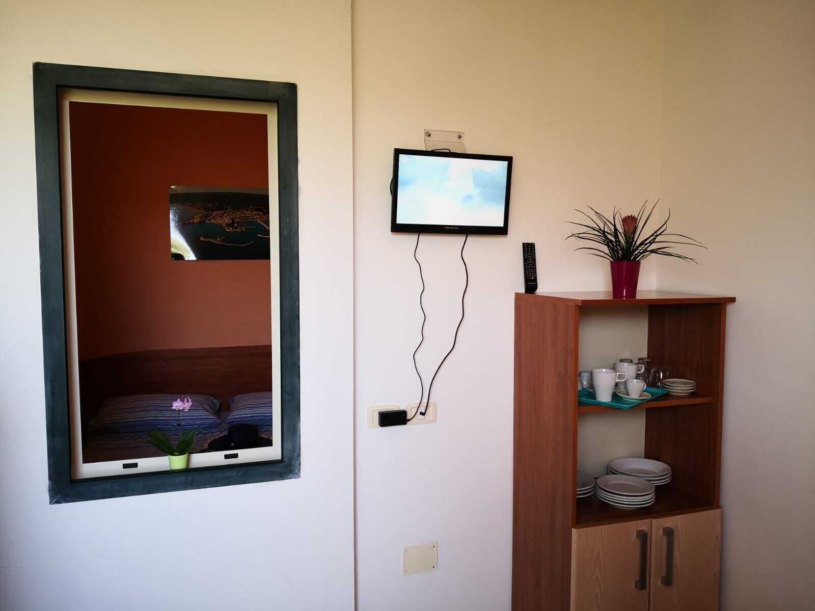 Quarto com janela, televisão fixada na parede, armário de madeira com louças e uma planta em vaso.