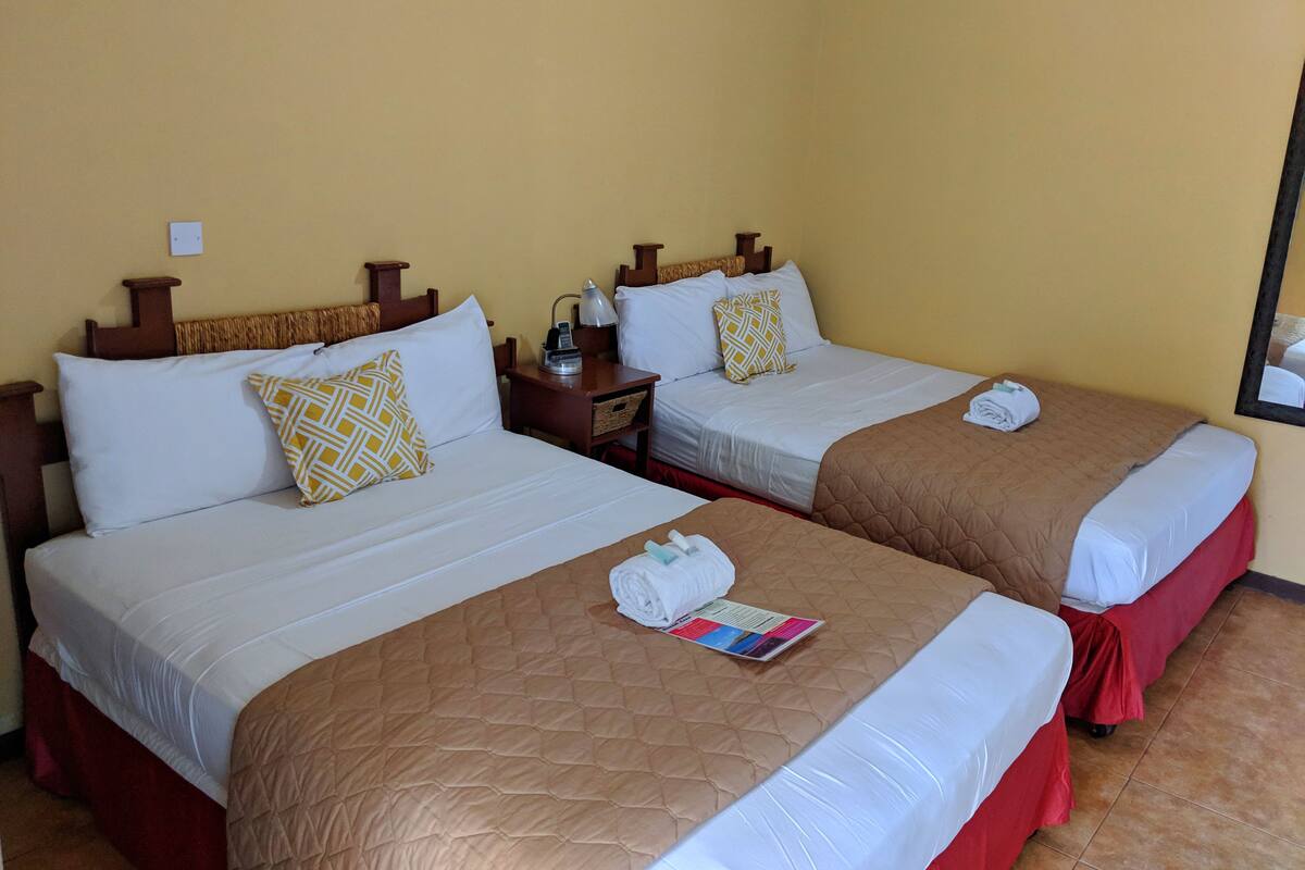 Duas camas de casal com roupa de cama branca e marrom, uma mesinha de cabeceira com luminária e uma cesta pequena em cada cama.
