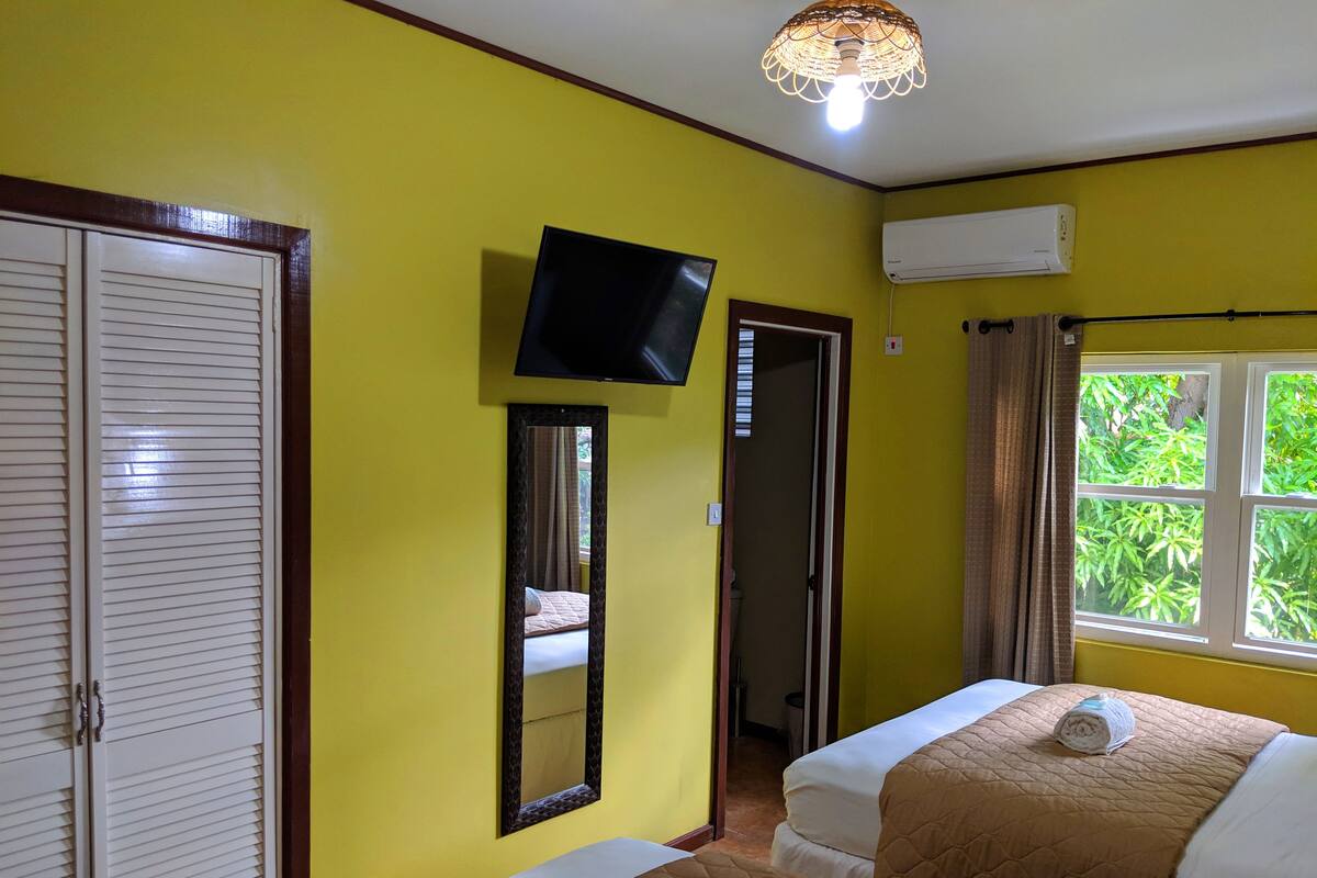 Quarto de hotel com paredes amarelas, uma cama com colcha marrom, televisão fixada na parede e ar condicionado.