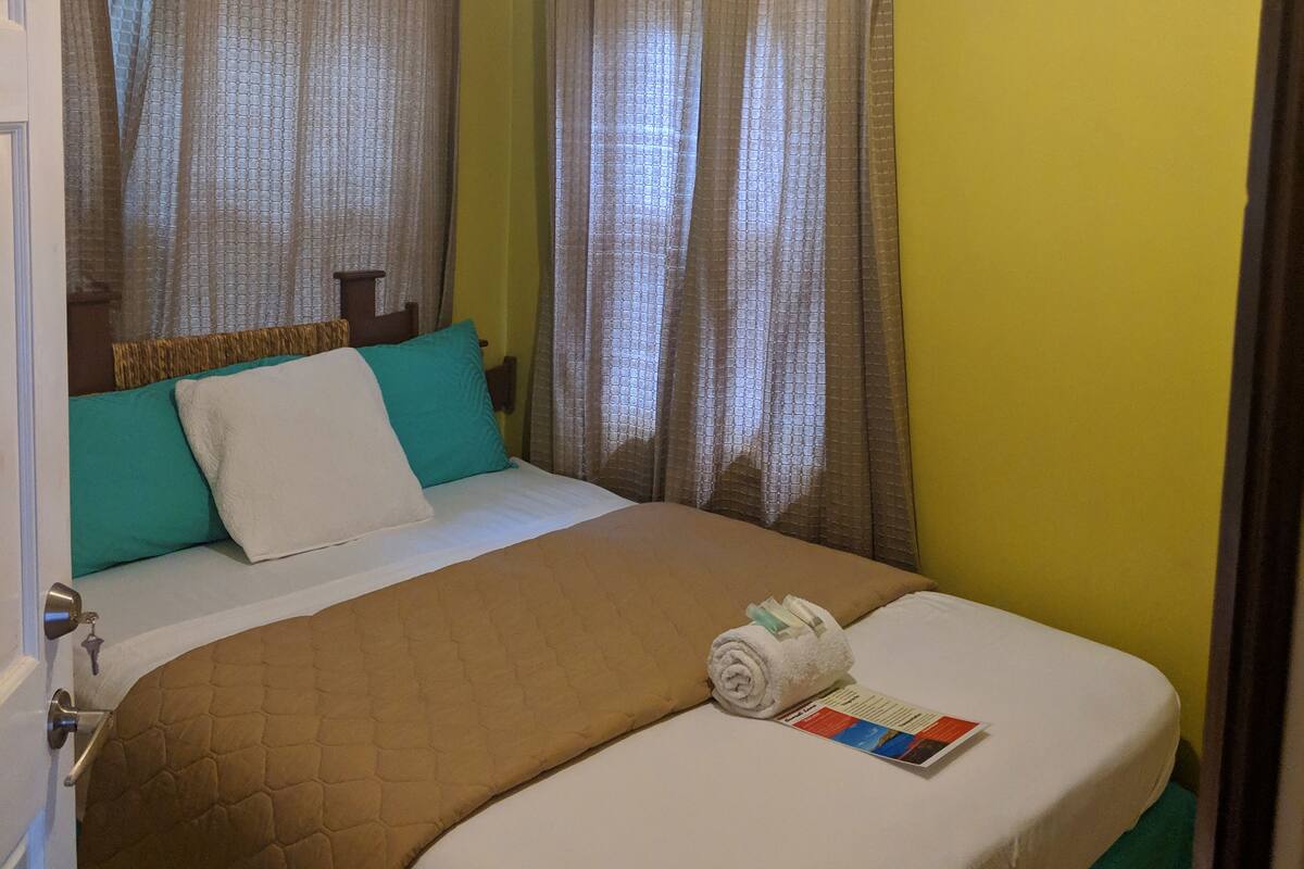 Cama bem arrumada com colcha marrom, travesseiro branco e travesseiro verde, cortinas estampadas e luminária acima.