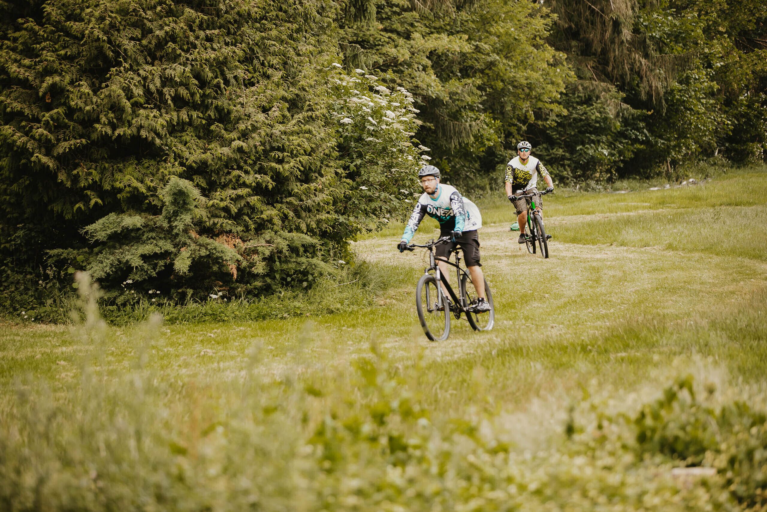 Dois ciclistas em uma trilha gramada, cercados por árvores densas.