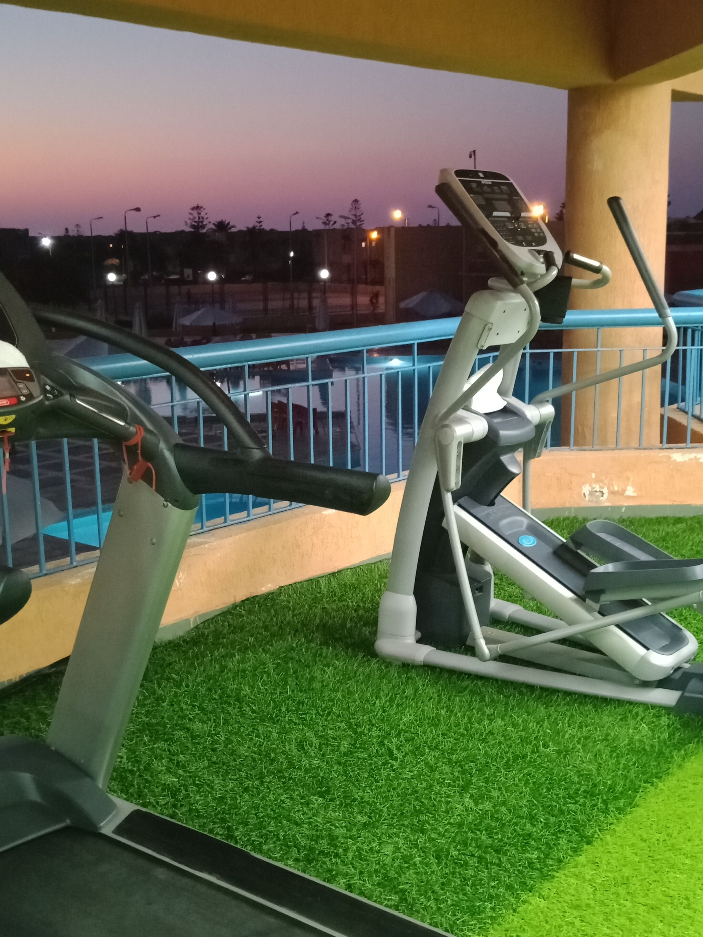 Uma academia ao ar livre com equipamentos de ginástica sobre grama sintética, cercada por um corrimão azul e uma estrutura de concreto.