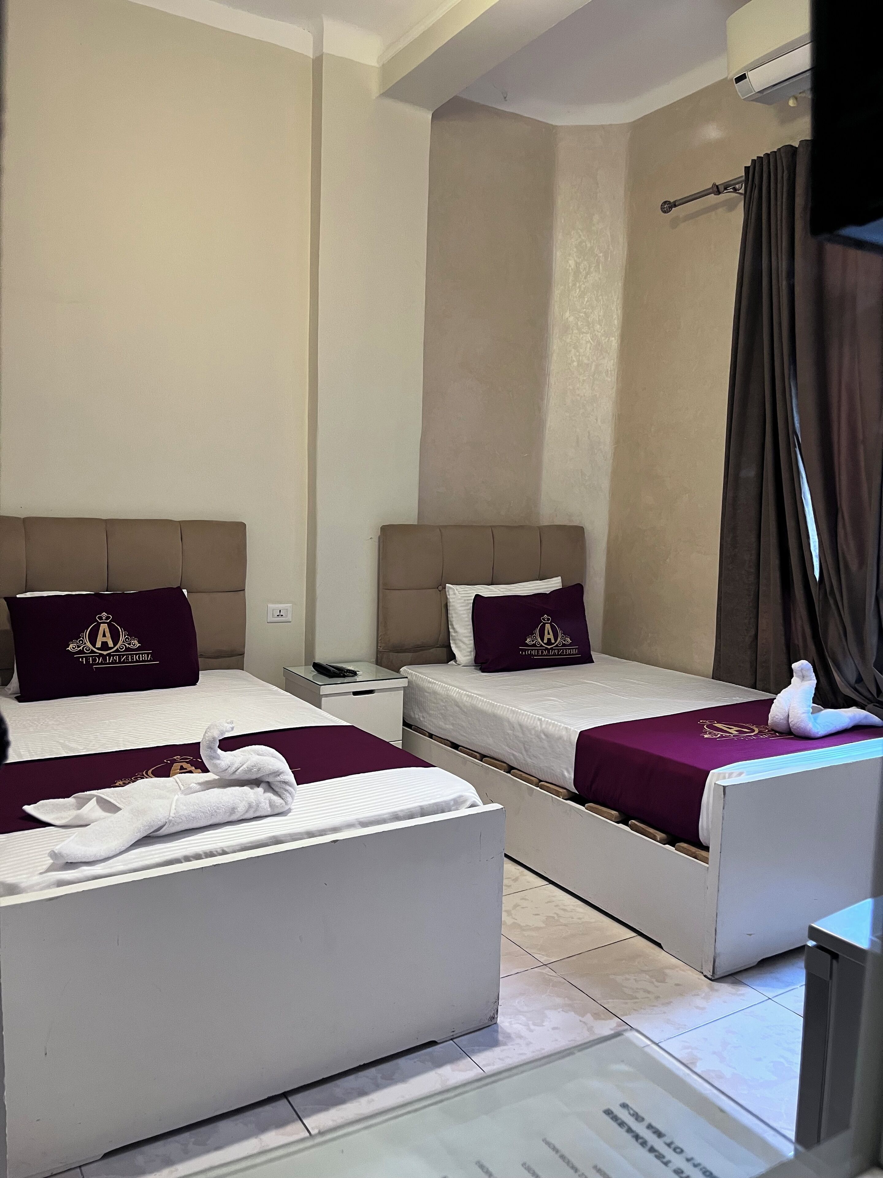 Quarto de hotel com duas camas, cada uma com cabeceira e travesseiro com monograma. Há um quadro branco com anotações, uma mesa de cabeceira com um controle remoto e uma toalha no chão.