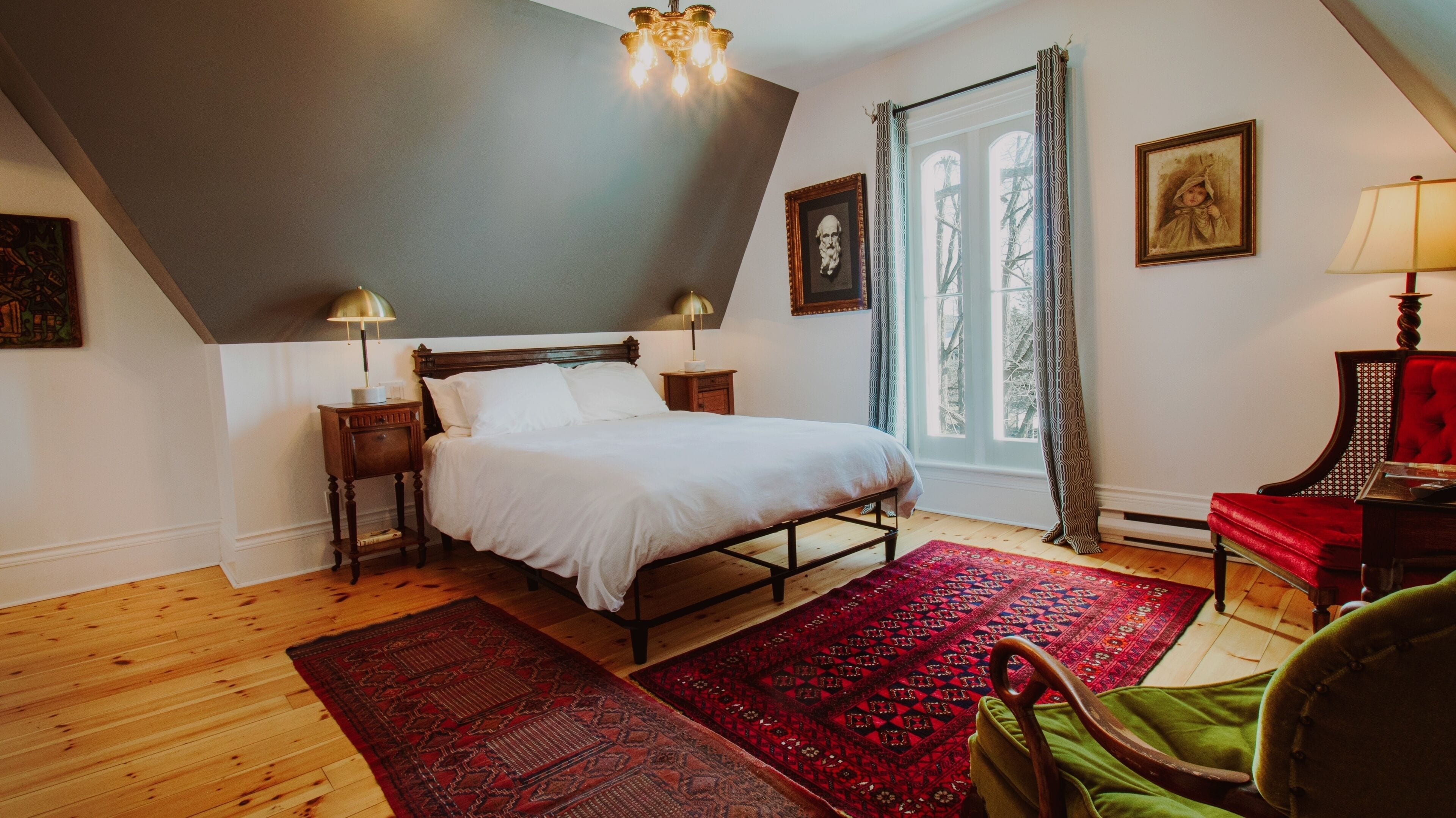 Quarto com cama, duas mesas de cabeceira, um candelabro, janela com cortinas, uma cadeira vermelha e um tapete estampado.