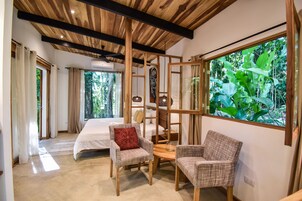 Quarto com cama, duas cadeiras, teto de madeira e grandes janelas com vista para o verde.