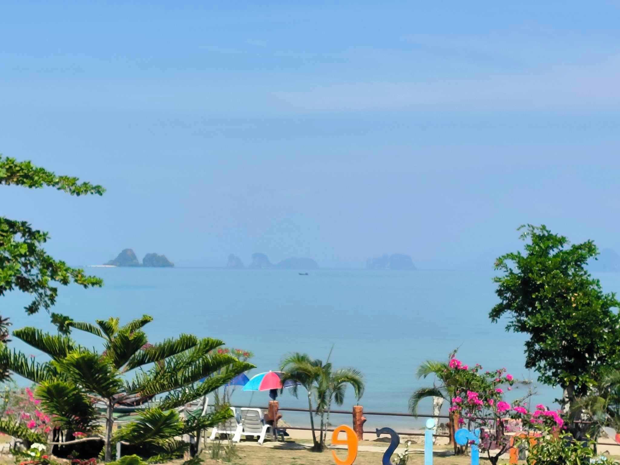 Vista da praia com palmeiras, uma cadeira e uma pessoa sob um guarda-sol colorido.