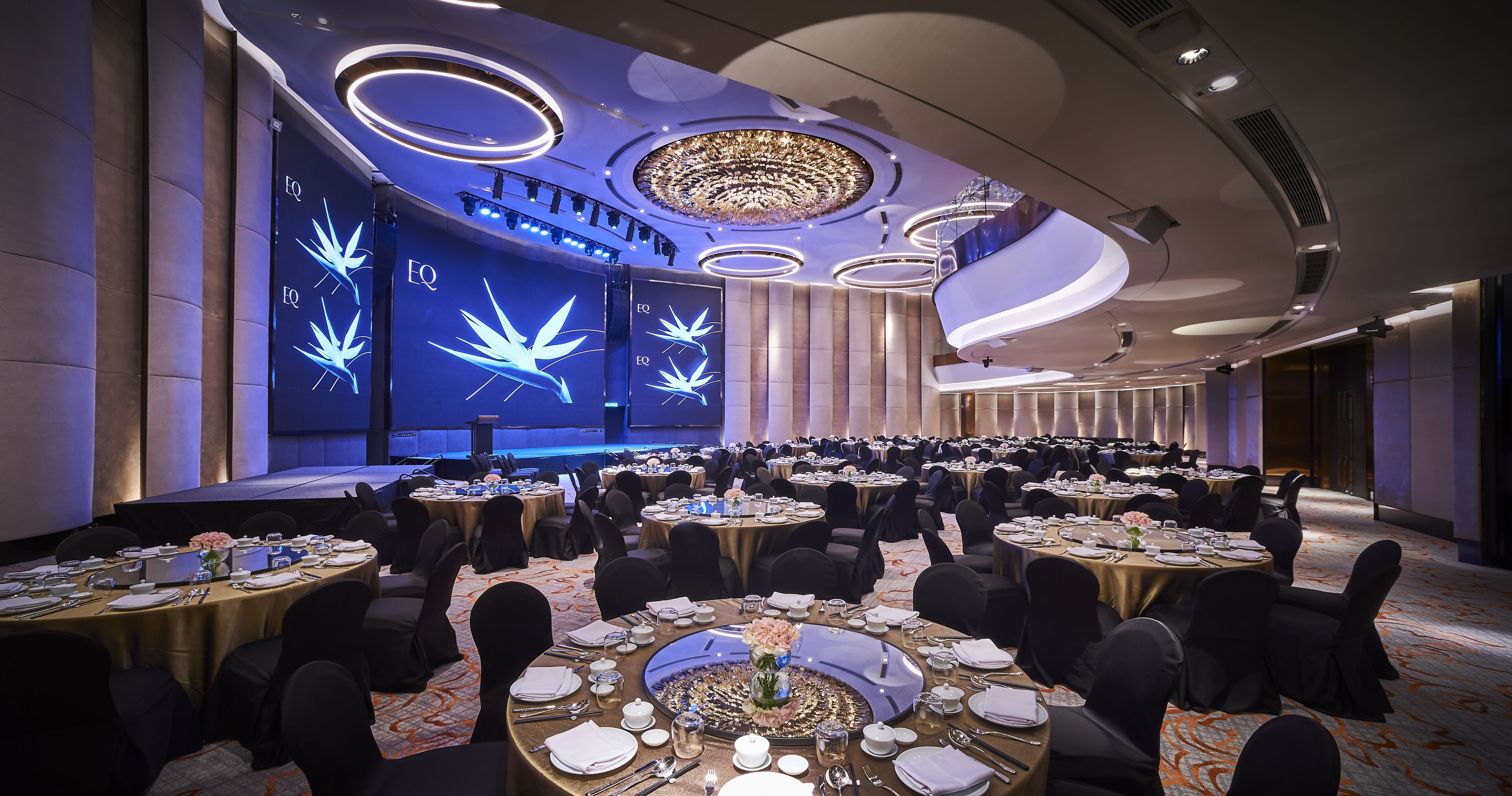 Um amplo salão de banquetes com mesas redondas dispostas para refeições, um palco com fundo azul e um lustre moderno.