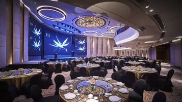 Um amplo salão de banquetes com mesas redondas dispostas para refeições, um palco com fundo azul e um lustre moderno.