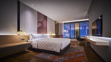 Quarto de hotel moderno com uma cama grande, uma escrivaninha, um banheiro com toucador e vista para a cidade à noite.