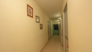 Um corredor estreito com paredes de cor clara, chão de azulejos e várias portas de um lado. Há três quadros pendurados na parede.