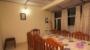 Uma área de refeições com uma mesa coberta por uma toalha florida, cercada por cadeiras de madeira. Um banquinho de madeira pequeno é visível à esquerda.