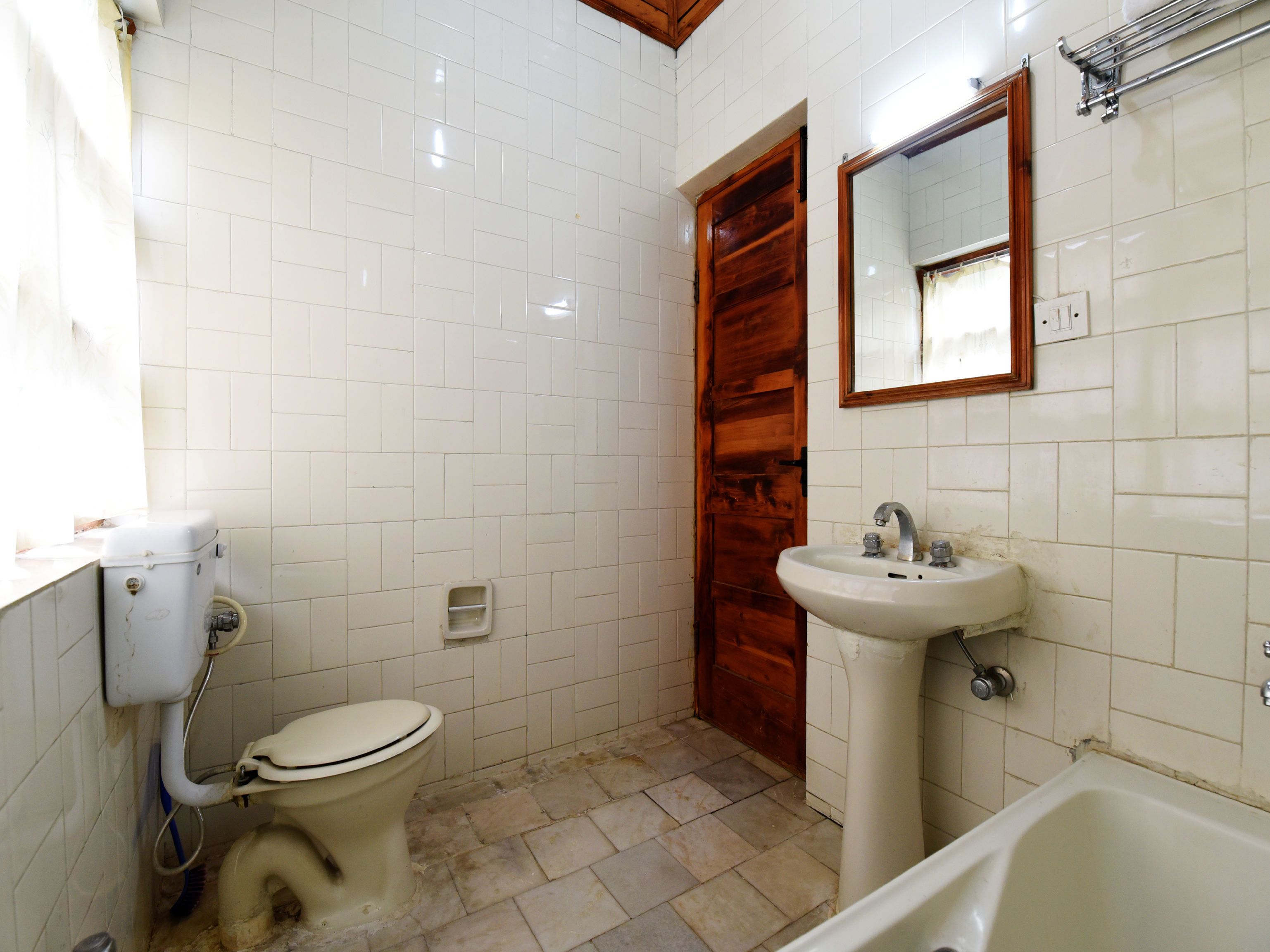Um banheiro com paredes em azulejos brancos, porta de madeira, vaso sanitário branco, pia e banheira.