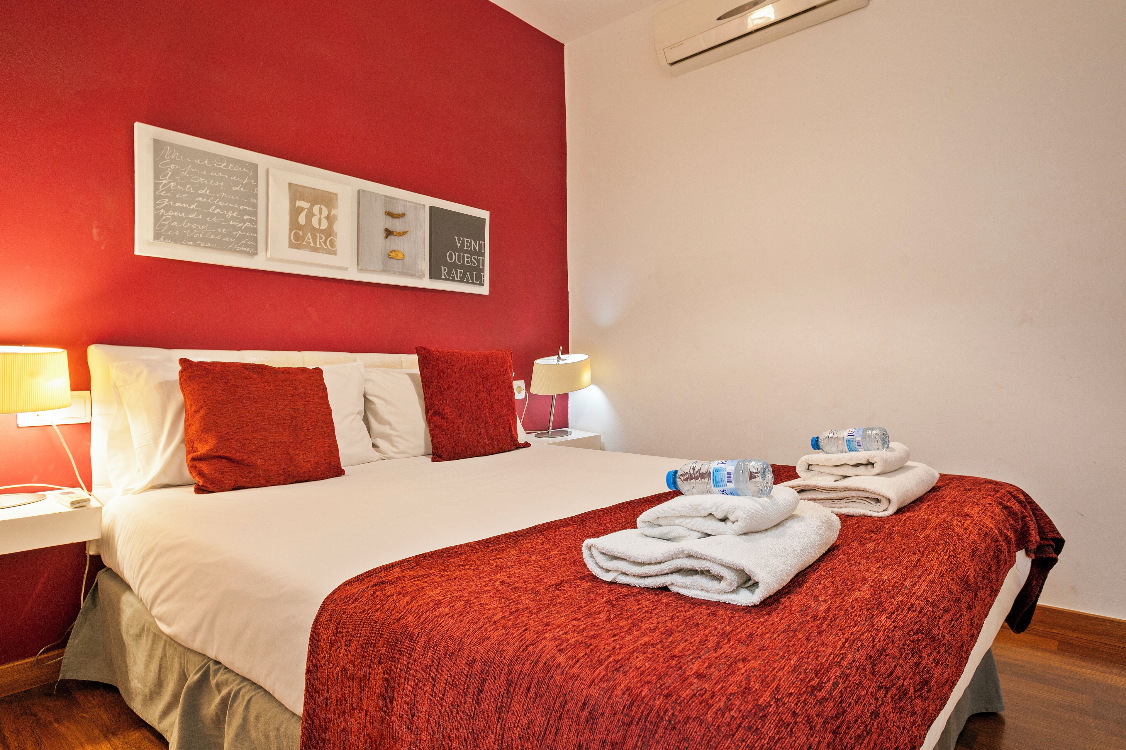 Quarto de hotel com uma parede em destaque vermelha, uma cama grande com roupa de cama branca, almofadas brancas e vermelhas e um cobertor vermelho. Há duas mesas de cabeceira com luminárias e uma garrafa de água na cama.