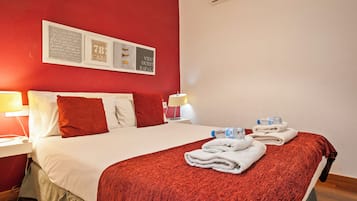 Quarto de hotel com uma parede em destaque vermelha, uma cama grande com roupa de cama branca, almofadas brancas e vermelhas e um cobertor vermelho. Há duas mesas de cabeceira com luminárias e uma garrafa de água na cama.