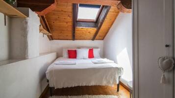 Quarto com teto inclinado, claraboia, cama com roupa de cama branca e almofadas vermelhas, e tapete branco no chão.