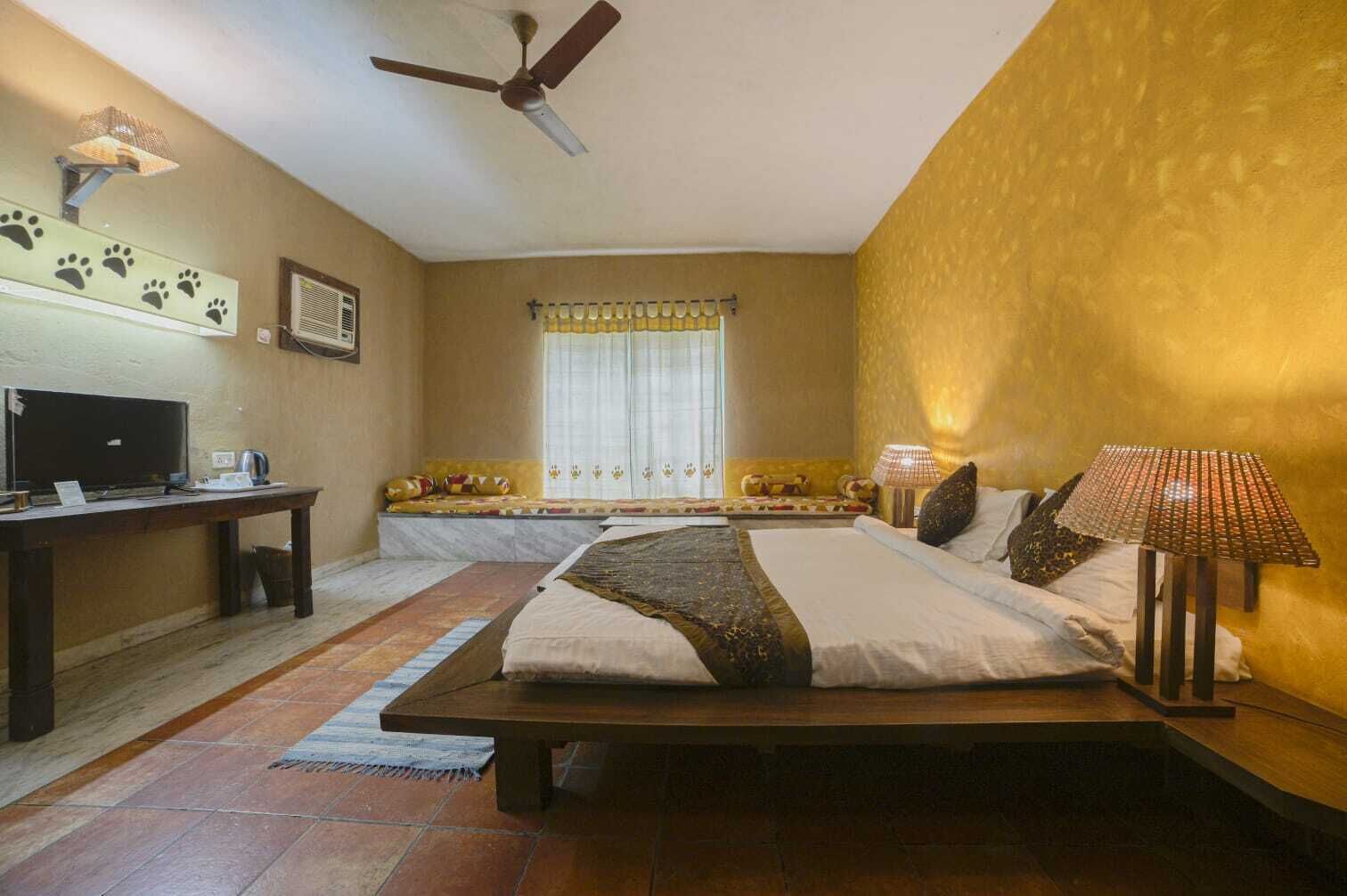 Quarto de hotel com duas camas, uma mesa de trabalho com televisão, uma janela com cortinas e um ventilador de teto.
