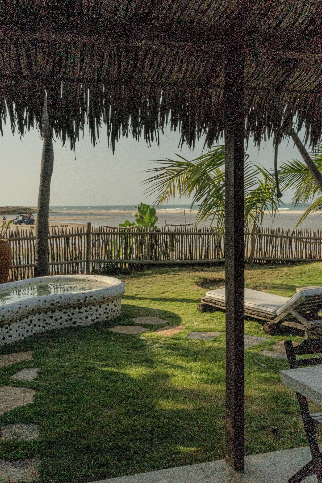Uma área externa coberta com piscina, cadeiras de descanso e vista para a praia.