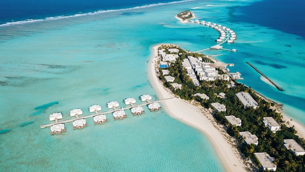 Vista aérea de uma ilha resort com bangalôs sobre a água, uma praia e águas turquesa cristalinas.
