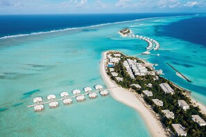 Vista aérea de uma ilha resort com bangalôs sobre a água, uma praia e águas turquesa cristalinas.