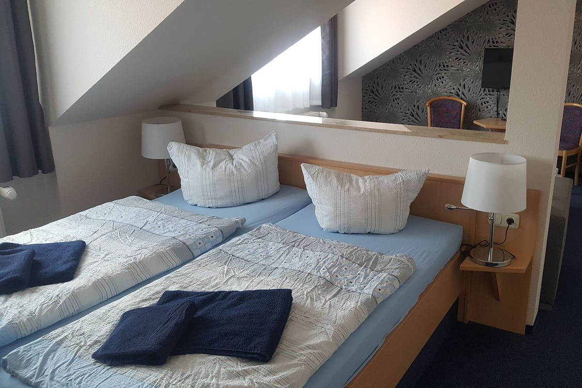 Quarto de hotel com duas camas, cada uma com travesseiro e cobertor. Há uma mesa de cabeceira com abajur, uma cabeceira de madeira e um espelho. O quarto possui um teto com design inclinado e uma janela com cortinas.