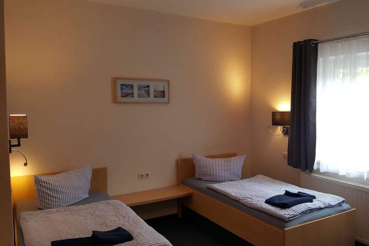 Quarto de hotel com duas camas de solteiro, uma pequena mesa de cabeceira, uma luminária de parede, uma janela com cortinas e um quadro pendurado na parede.