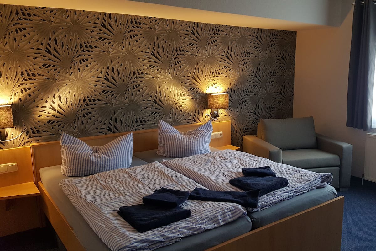 Quarto de hotel com uma cama grande, duas mesas de cabeceira, uma cadeira, um painel decorativo na parede e uma janela com cortinas.