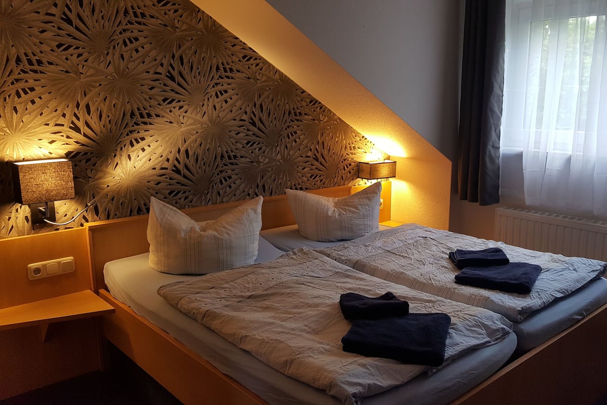 Quarto de hotel com cama, duas luminárias de cabeceira, um painel decorativo na parede e uma janela com cortinas.