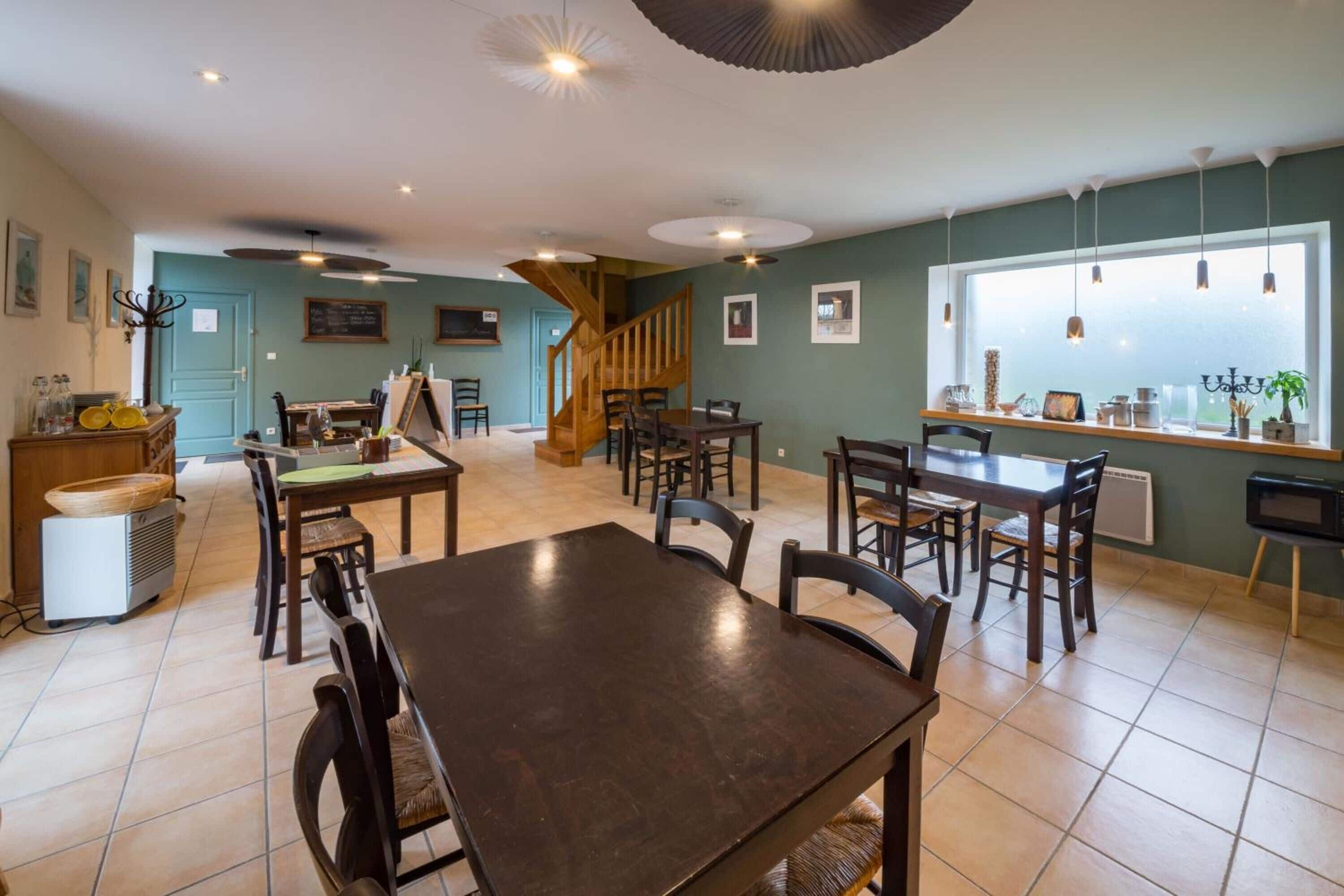 Uma sala de jantar ampla, com uma grande mesa de madeira e várias cadeiras, área de cozinha com janela e escada que leva a outro andar.