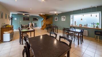 Uma sala de jantar ampla, com uma grande mesa de madeira e várias cadeiras, área de cozinha com janela e escada que leva a outro andar.