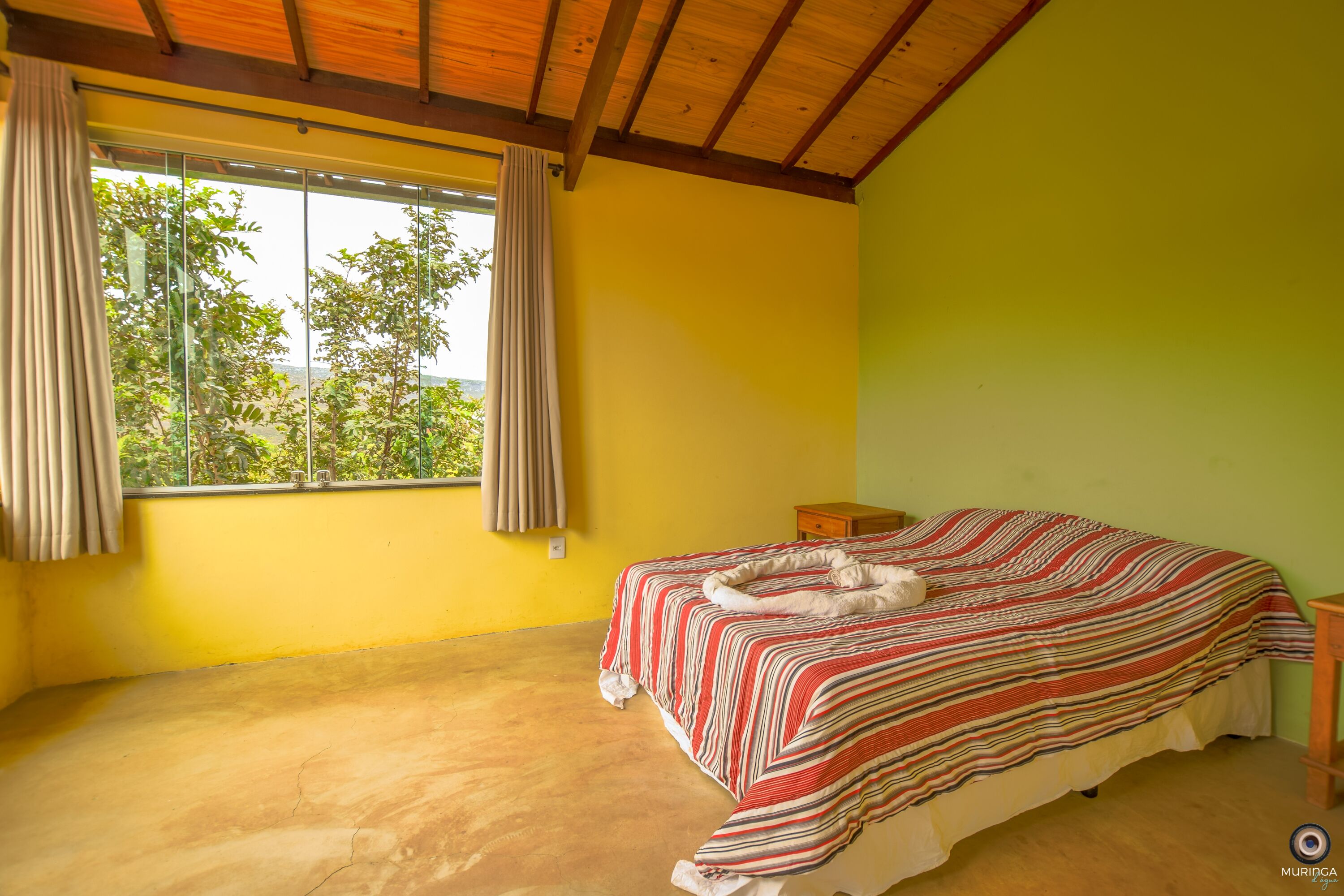 Quarto com cama, janela e vista para o verde lá fora.