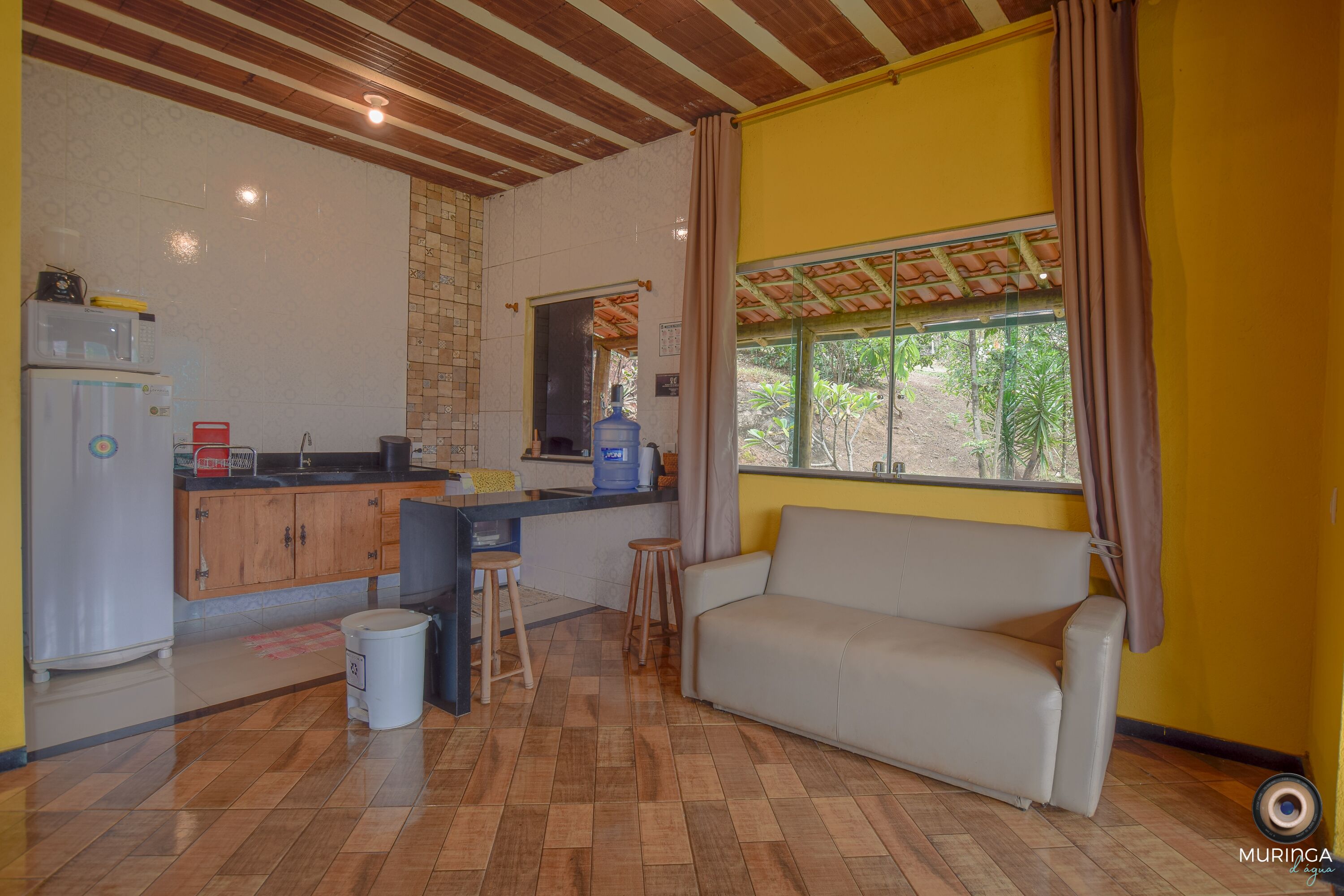 Uma cozinha compacta com geladeira, micro-ondas e pia. Há um sofá bege e uma janela com cortinas.