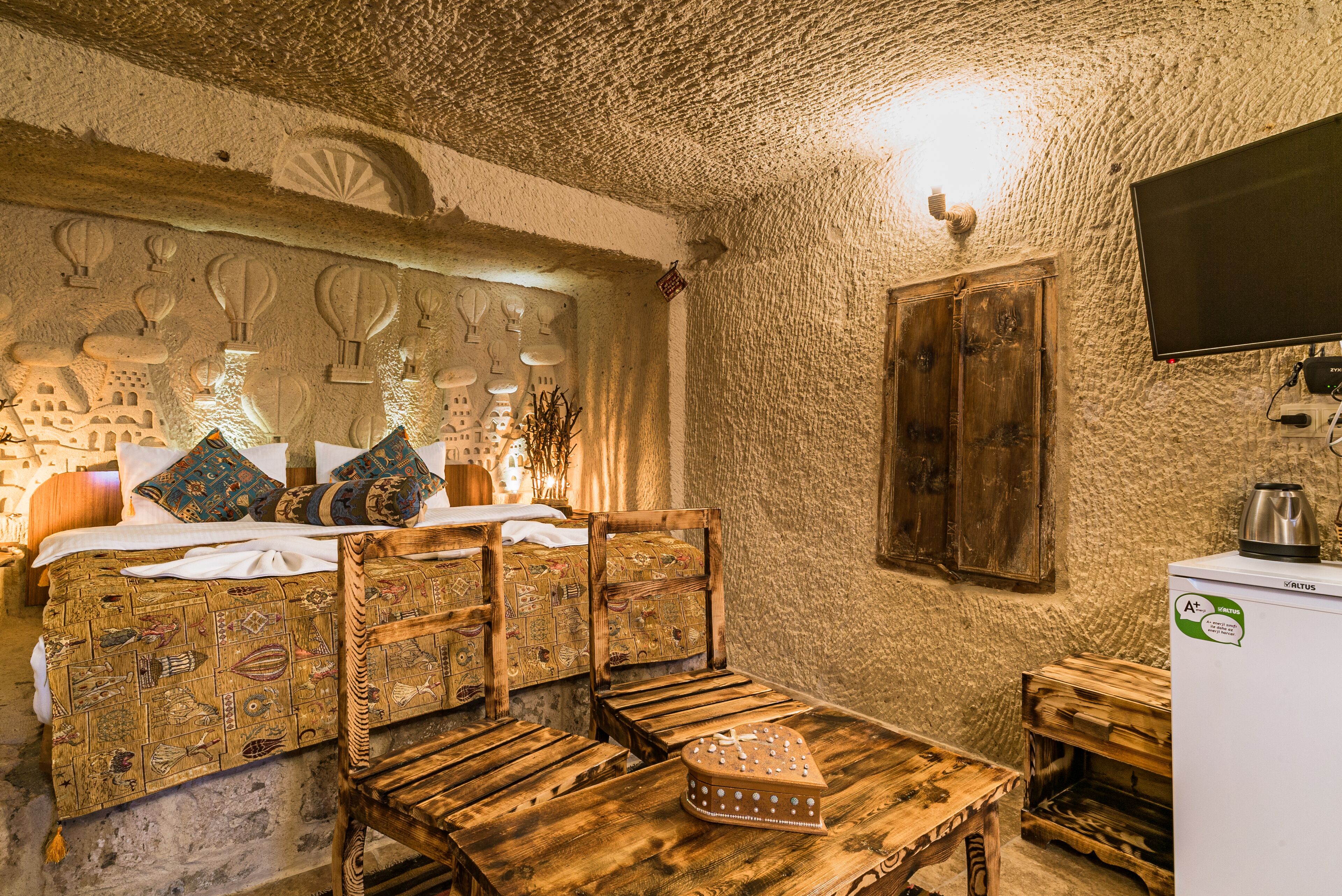 Quarto com cama, cadeiras de madeira, mesa, televisão e frigorífico.