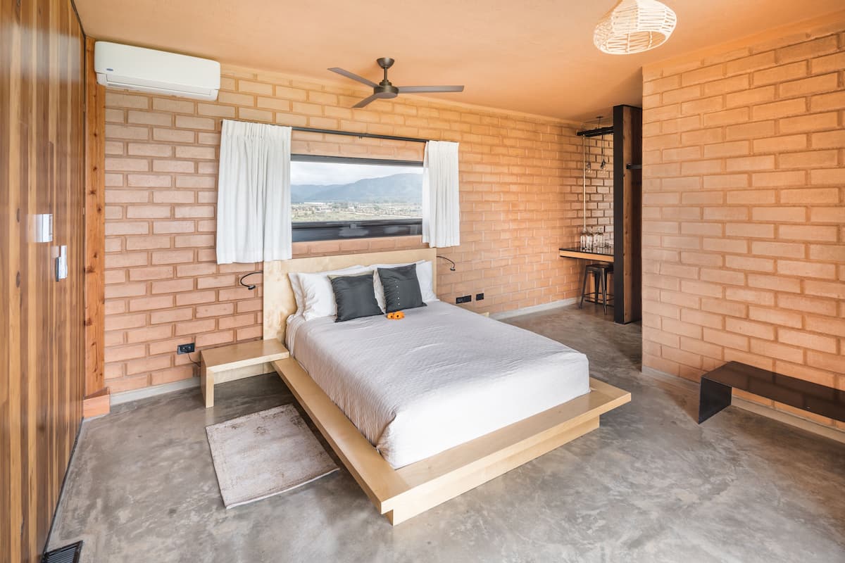 Quarto com parede de tijolos, cama com estrutura de madeira, ventilador de teto e vista para a cidade através de uma janela.