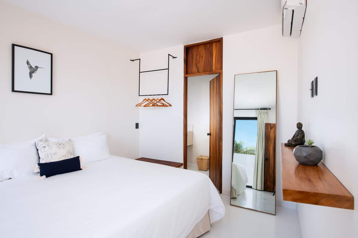 Quarto moderno com cama grande, prateleira de madeira, espelho e luminária pendente.