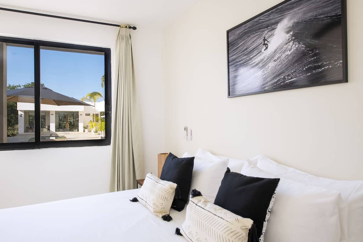 Quarto de hotel com cama, uma janela ampla, um quadro de um surfista e vista para um edifício.