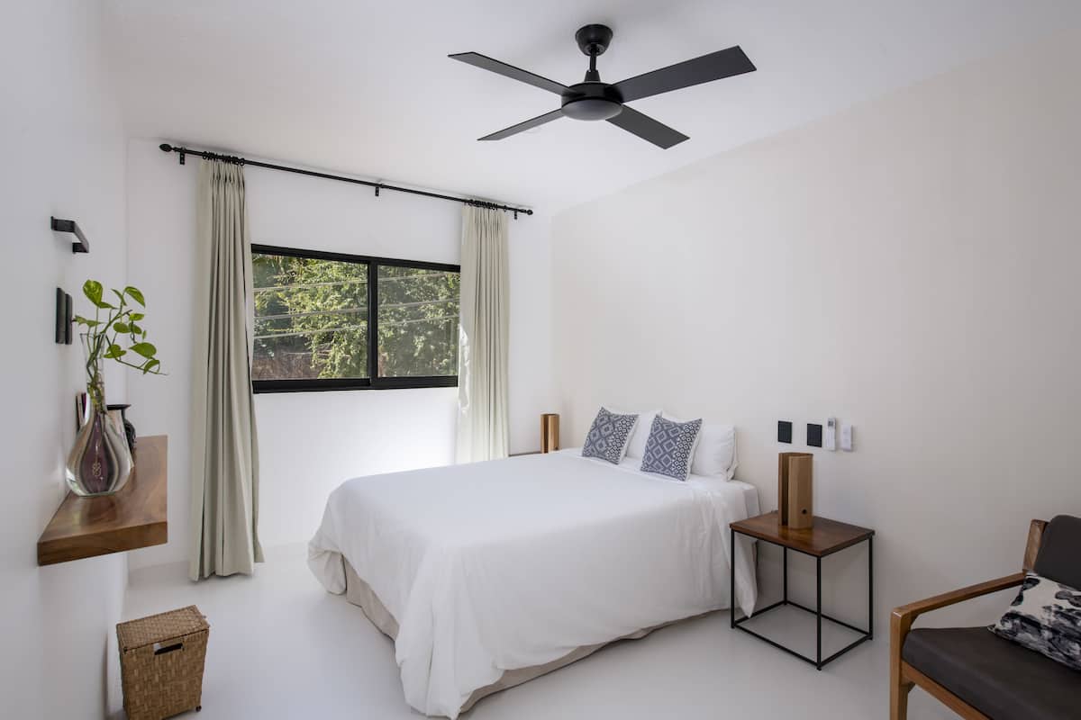 Quarto moderno com cama grande, ventilador de teto, janela com cortinas, mesa de cabeceira e uma cadeira.