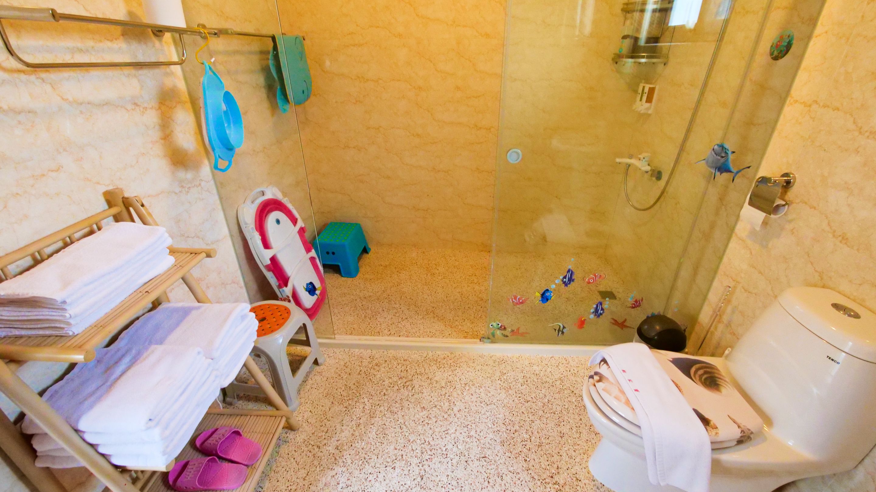 Casa de banho com cabine de chuveiro em vidro, sanita, lavatório e prateleira com toalhas e chinelos.