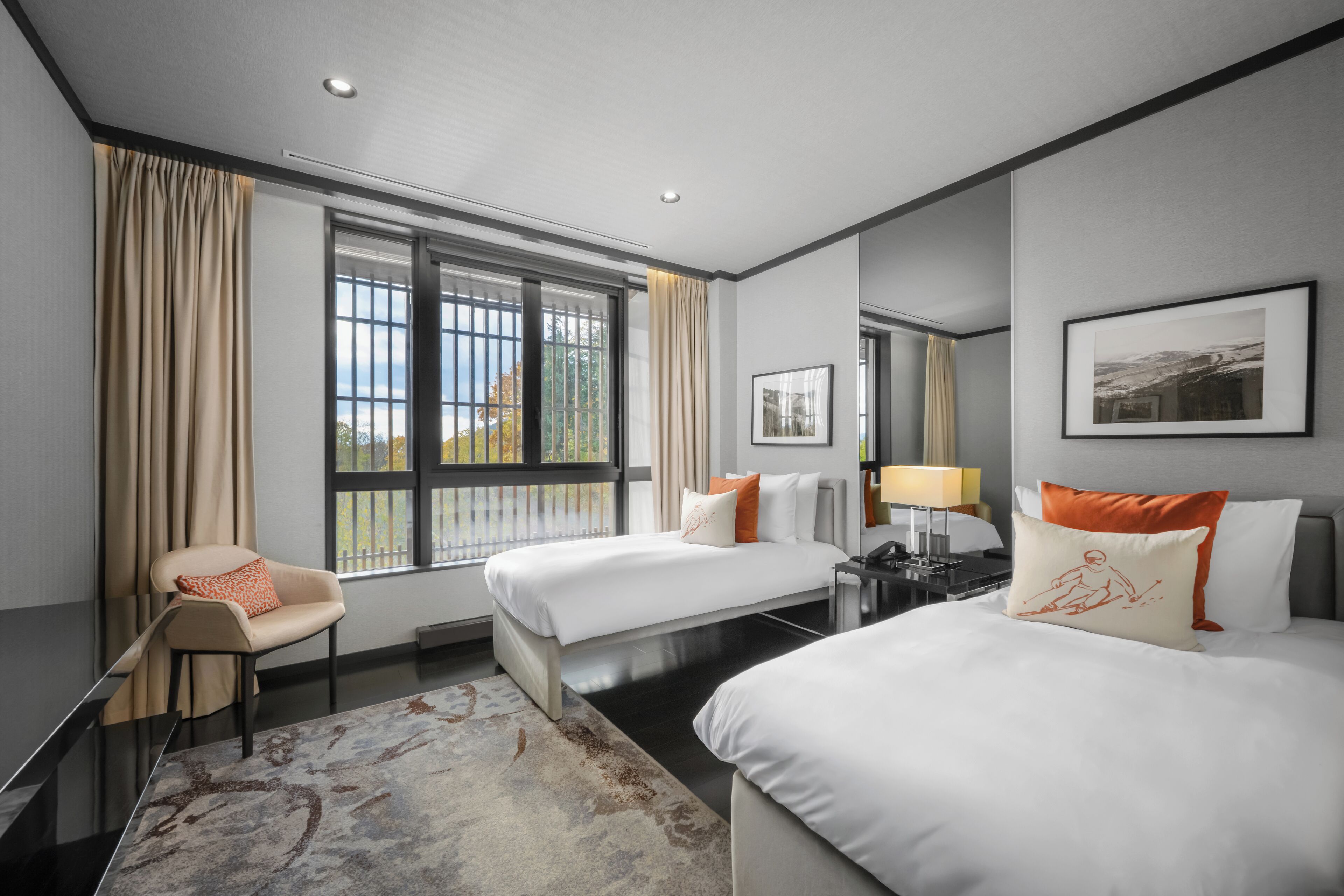 Quarto de hotel com duas camas, uma cadeira, uma escrivaninha, um abajur e uma janela com cortinas.