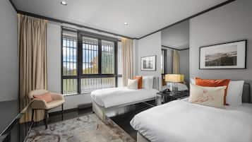 Quarto de hotel com duas camas, uma cadeira, uma escrivaninha, um abajur e uma janela com cortinas.