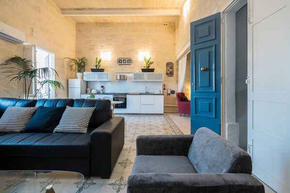 Sala de estar com sofá e poltrona, cozinha ao fundo, e uma porta azul.