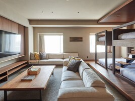 Quarto moderno e bem iluminado, com sofá, televisão, mesa de madeira e beliche.