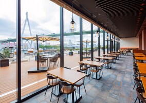 Restaurante com mesas ao ar livre, amplas janelas e vista para uma ponte.