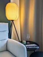 Um quarto com uma luminária, uma taça de vinho, uma vela e uma revista sobre uma mesa pequena.
