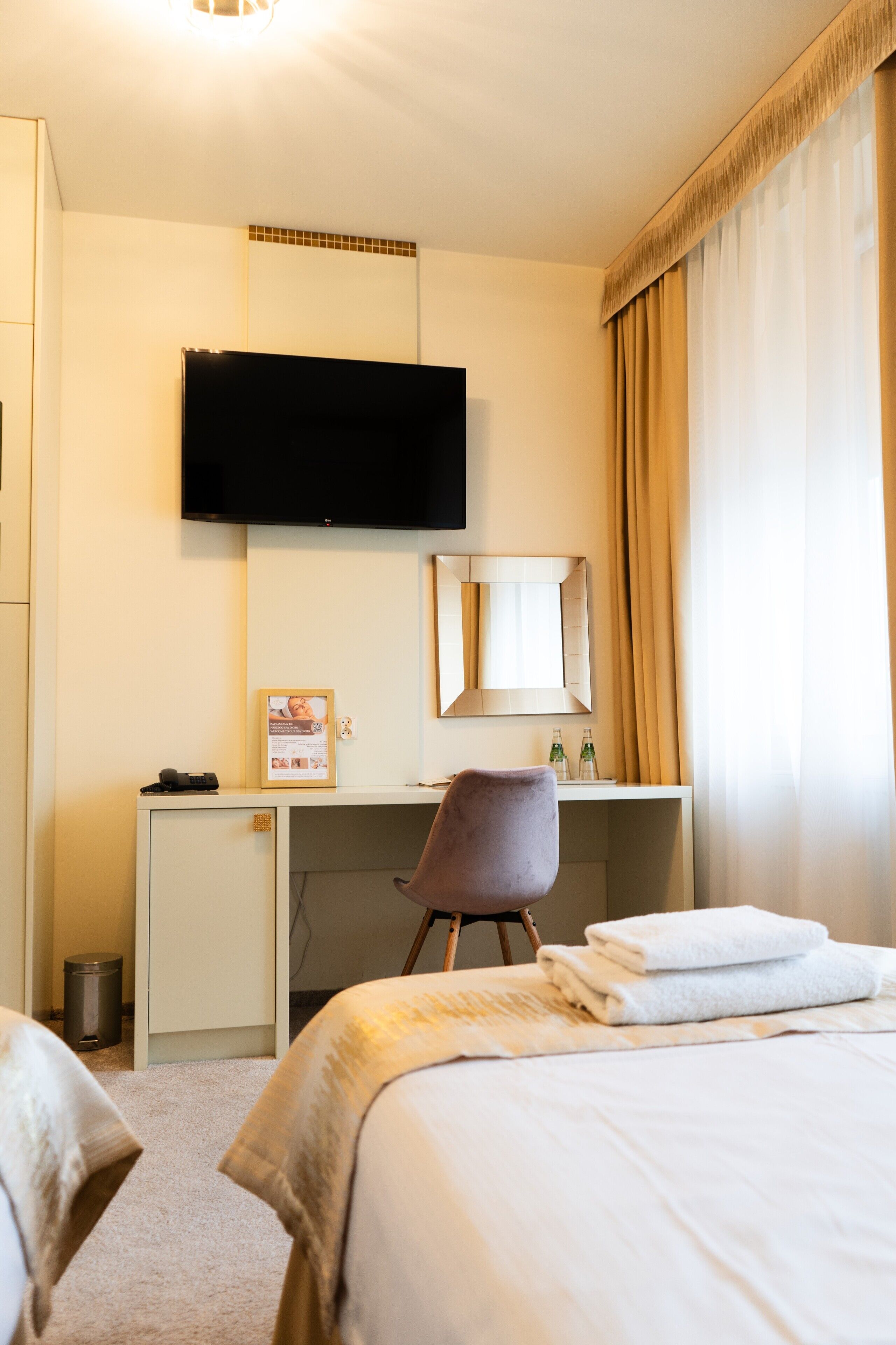 Quarto de hotel com uma cama, uma escrivaninha, uma cadeira, uma televisão na parede e uma janela com cortinas.