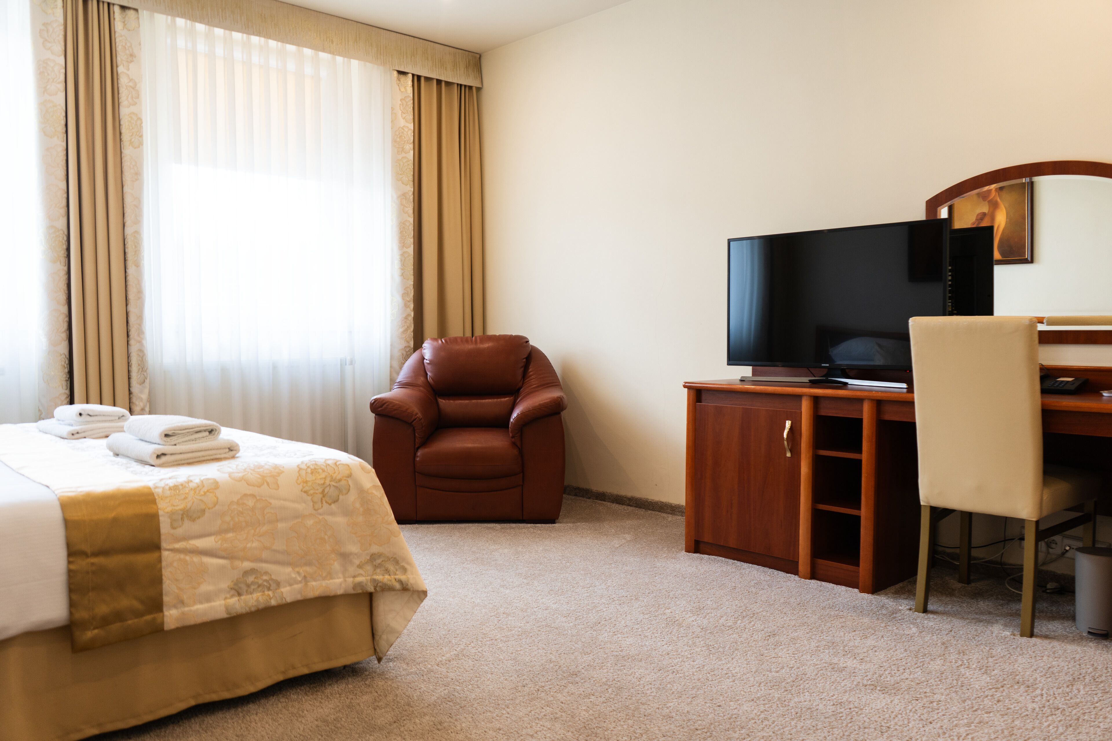 Quarto de hotel com cama, uma poltrona de couro marrom, escrivaninha com televisão e armário de madeira.