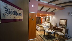 Quarto de hotel com cama, escrivaninha, cadeira e mesa com toalha. Há um quadro na parede e uma placa com a palavra 'Beethoven'.