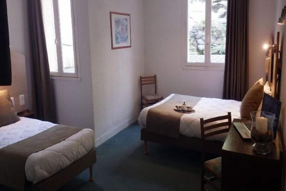 Quarto com duas camas, uma mesinha com um laptop, uma cadeira e uma janela com cortinas.